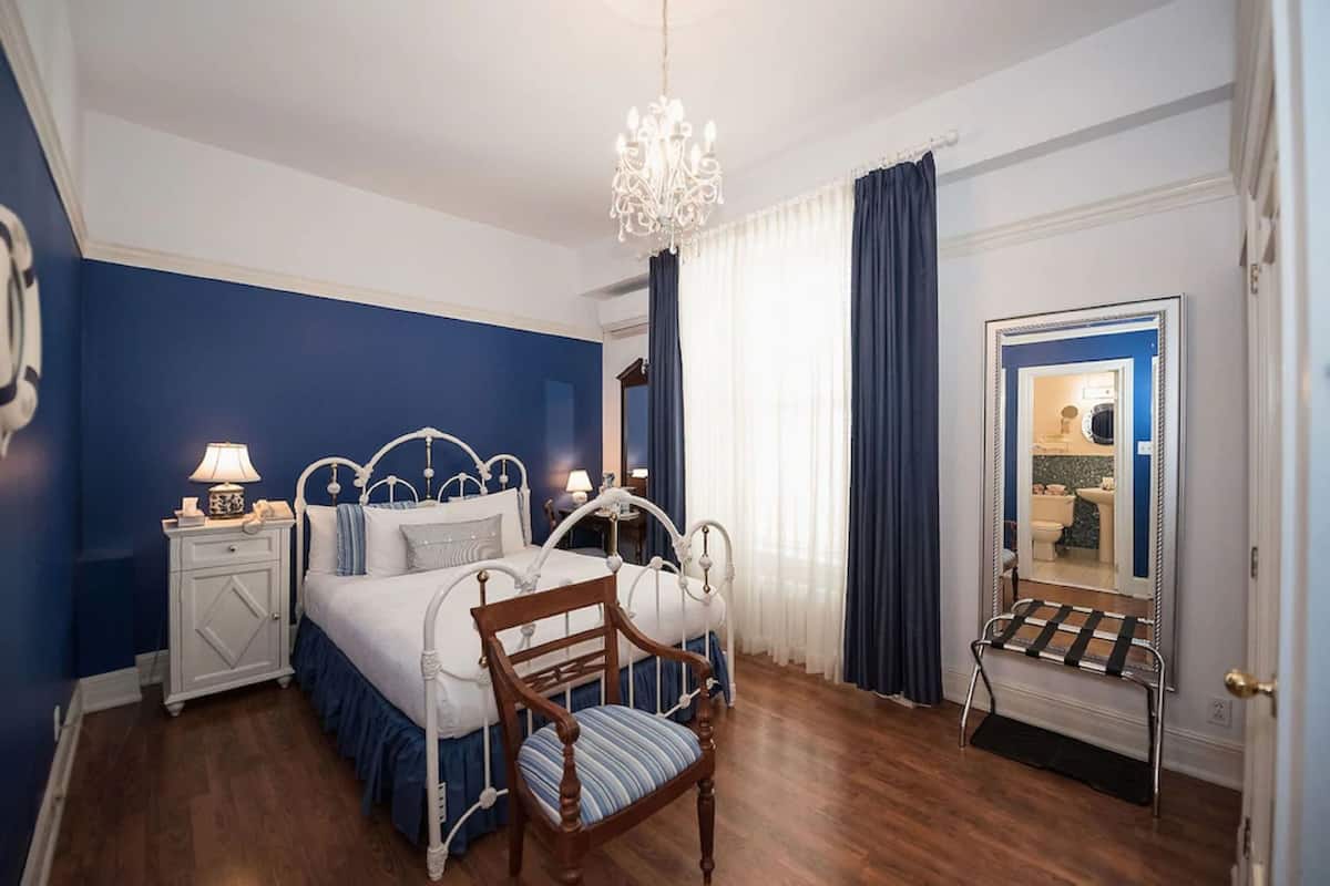 Quarto com estrutura de cama em metal branco, uma cadeira de madeira, mesa de cabeceira com luminária e um candelabro.