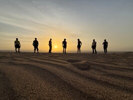 Um grupo de pessoas em uma superfície arenosa com o pôr do sol ao fundo.