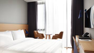 Quarto de hotel com uma cama grande, duas cadeiras, uma mesa pequena, uma escrivaninha com telefone e uma televisão.