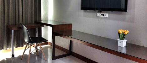 Quarto de hotel moderno com televisão de tela plana na parede, escrivaninha de madeira, cadeira preta e uma pequena planta em vaso.