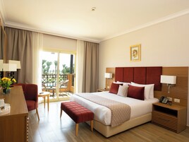 Quarto de hotel com cama, mesas de cabeceira, escrivaninha, cadeira e varanda com vista para palmeiras.