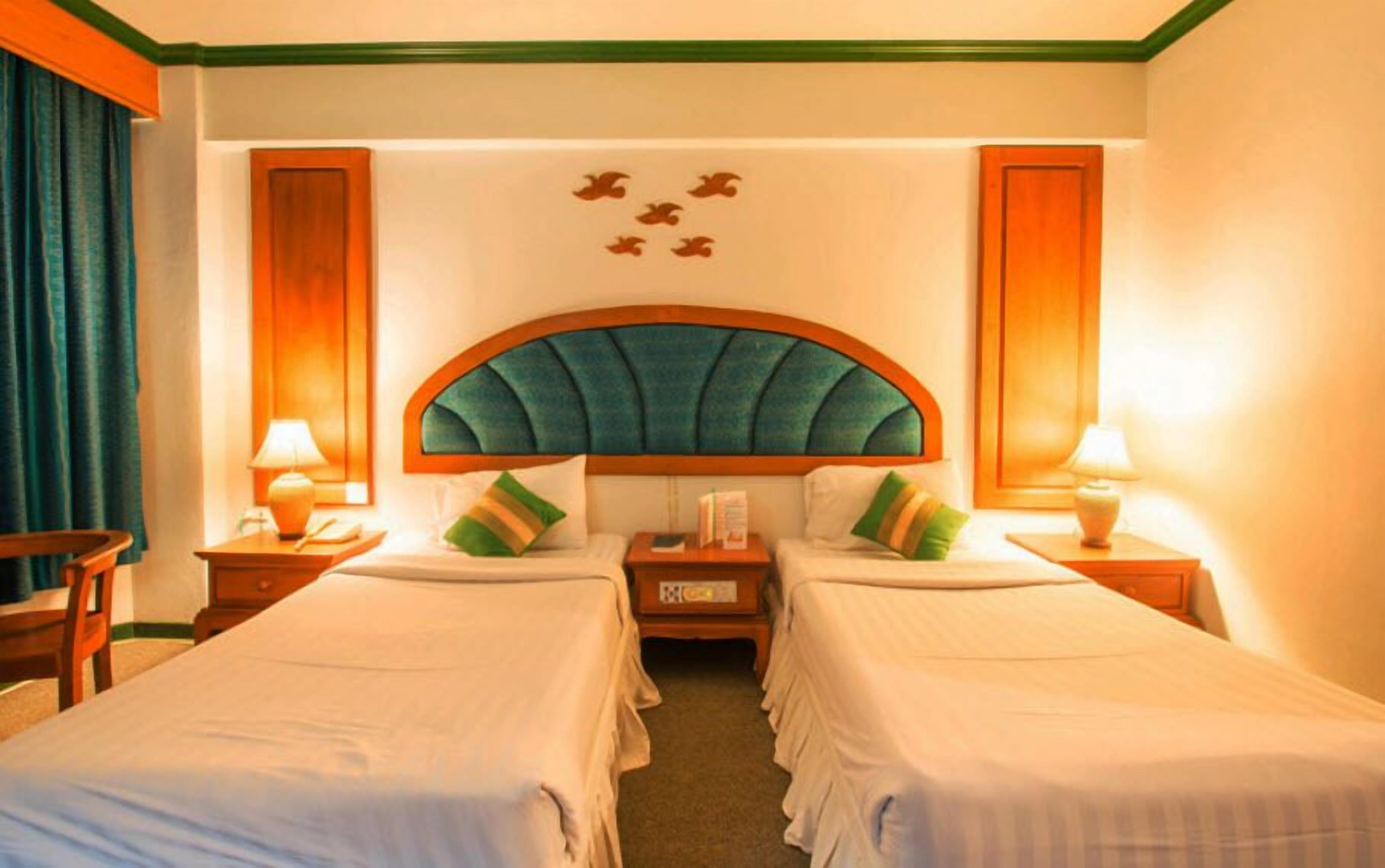 Quarto de hotel com duas camas, cabeceira de madeira, luminárias, mesa de cabeceira e um enfeite na parede.