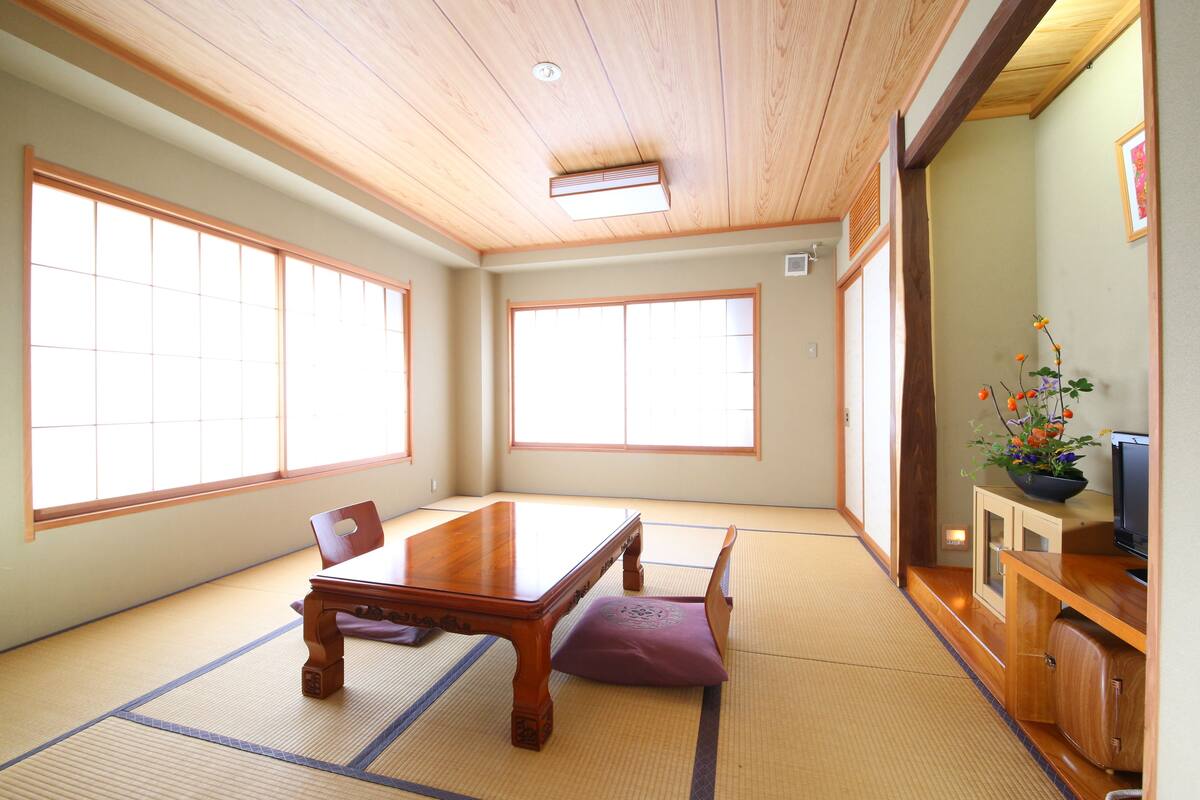 Um quarto japonês tradicional com tatames, uma mesa de madeira e uma televisão sobre um armário de madeira.