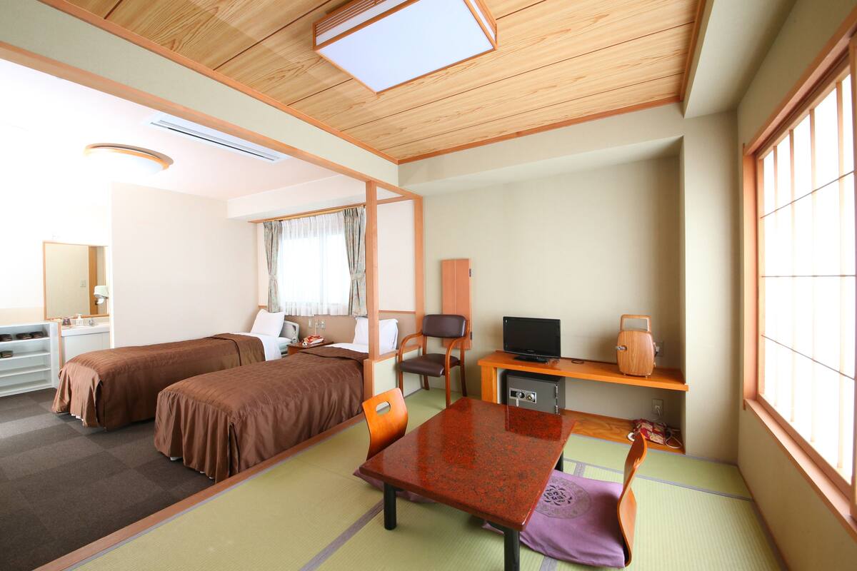 Quarto tradicional em estilo japonês, com piso de tatame, duas camas, uma escrivaninha com televisão e uma janela que permite a entrada de luz natural.