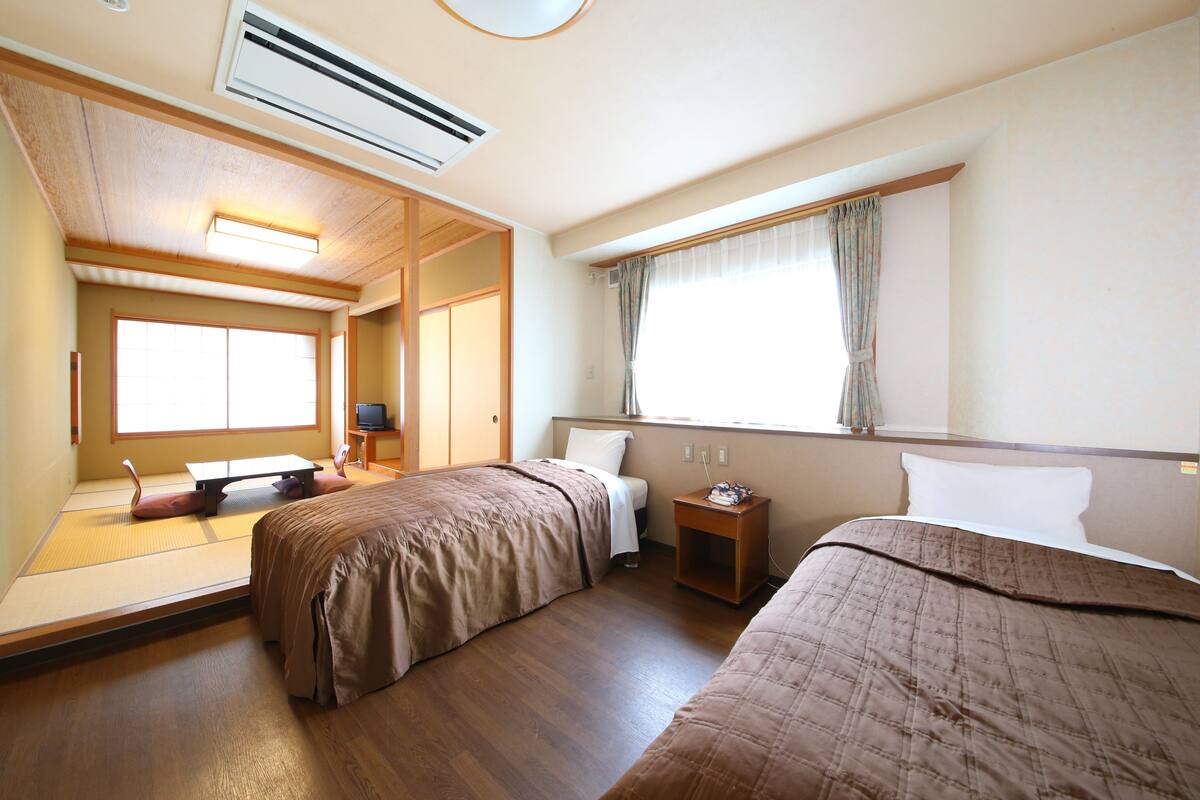Quarto de hotel com duas camas, uma mesinha, televisão e janela com cortinas.