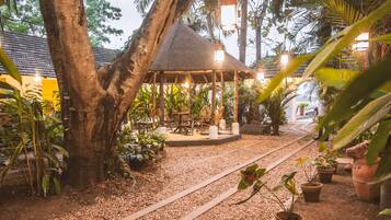 Uma trilha com um gazebo de colmo, rodeado por plantas em vasos e uma área de cascalho.