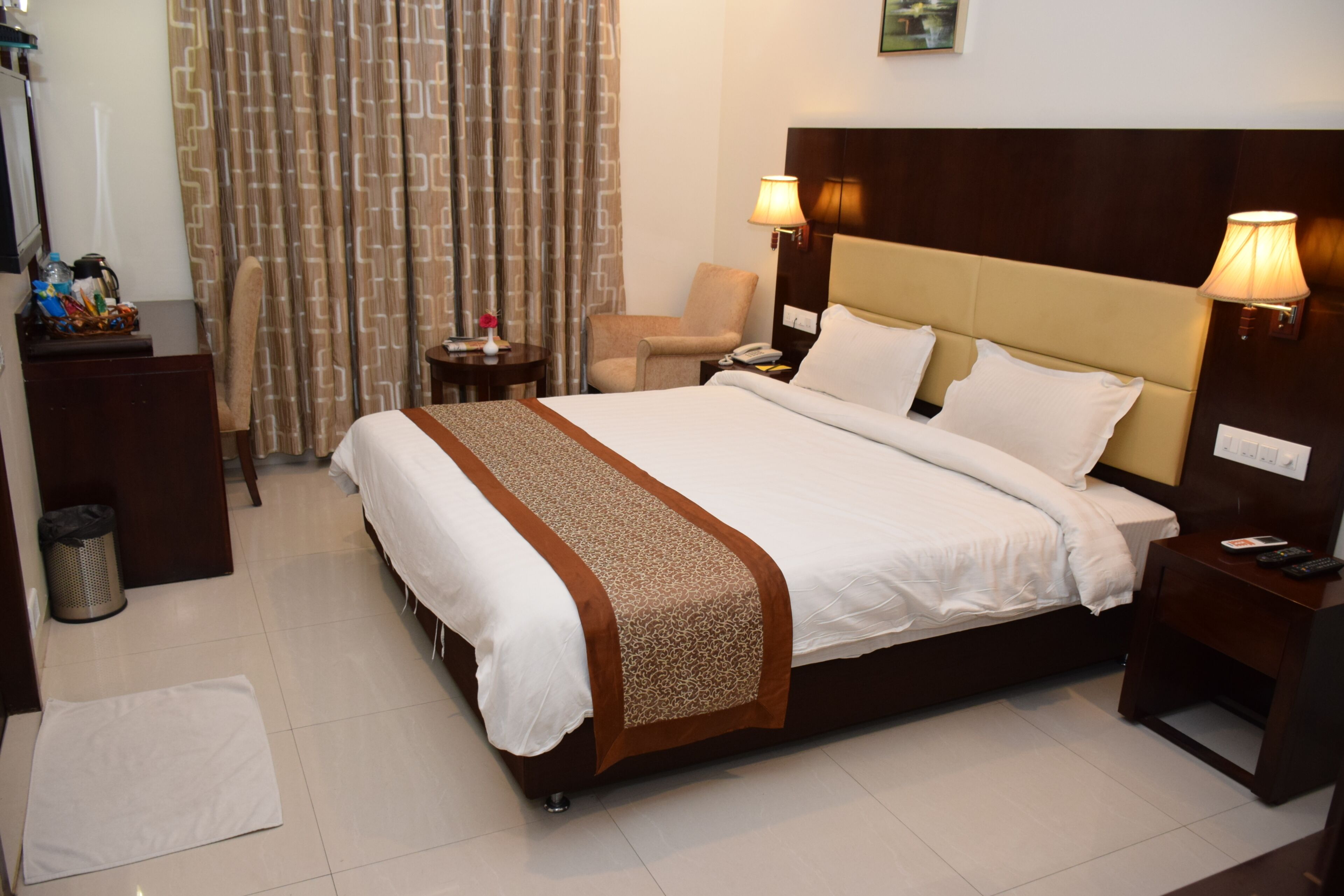 Quarto de hotel com uma cama grande, duas luminárias de cabeceira, uma cadeira, uma mesinha e um cesto de lixo.
