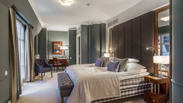 Quarto de hotel moderno com uma cama grande, uma escrivaninha com abajur, uma cadeira e vista para a cidade pela janela.