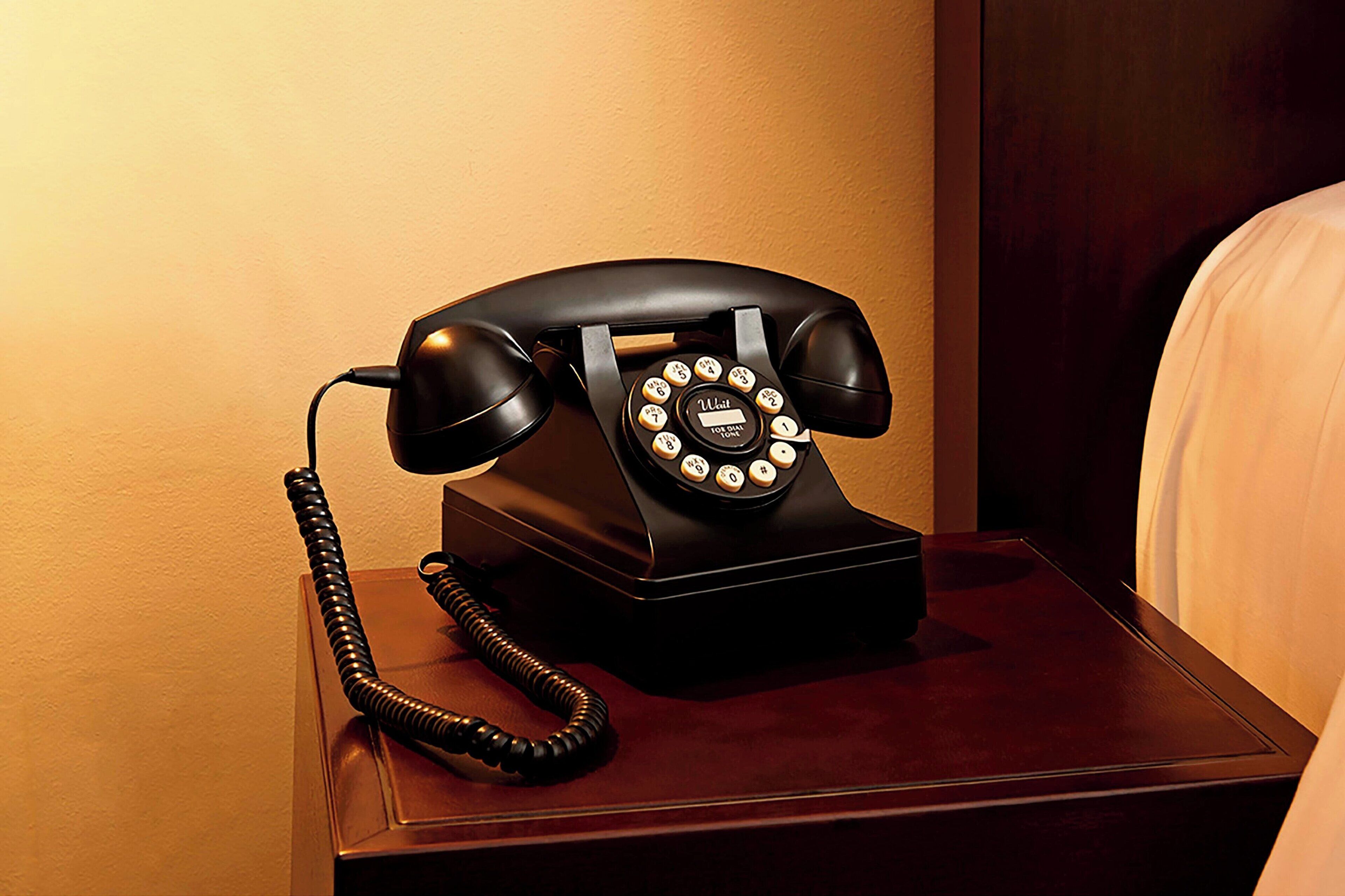 Um telefone antigo com discagem rotativa preta sobre uma mesa de cabeceira de madeira em quarto de hotel.