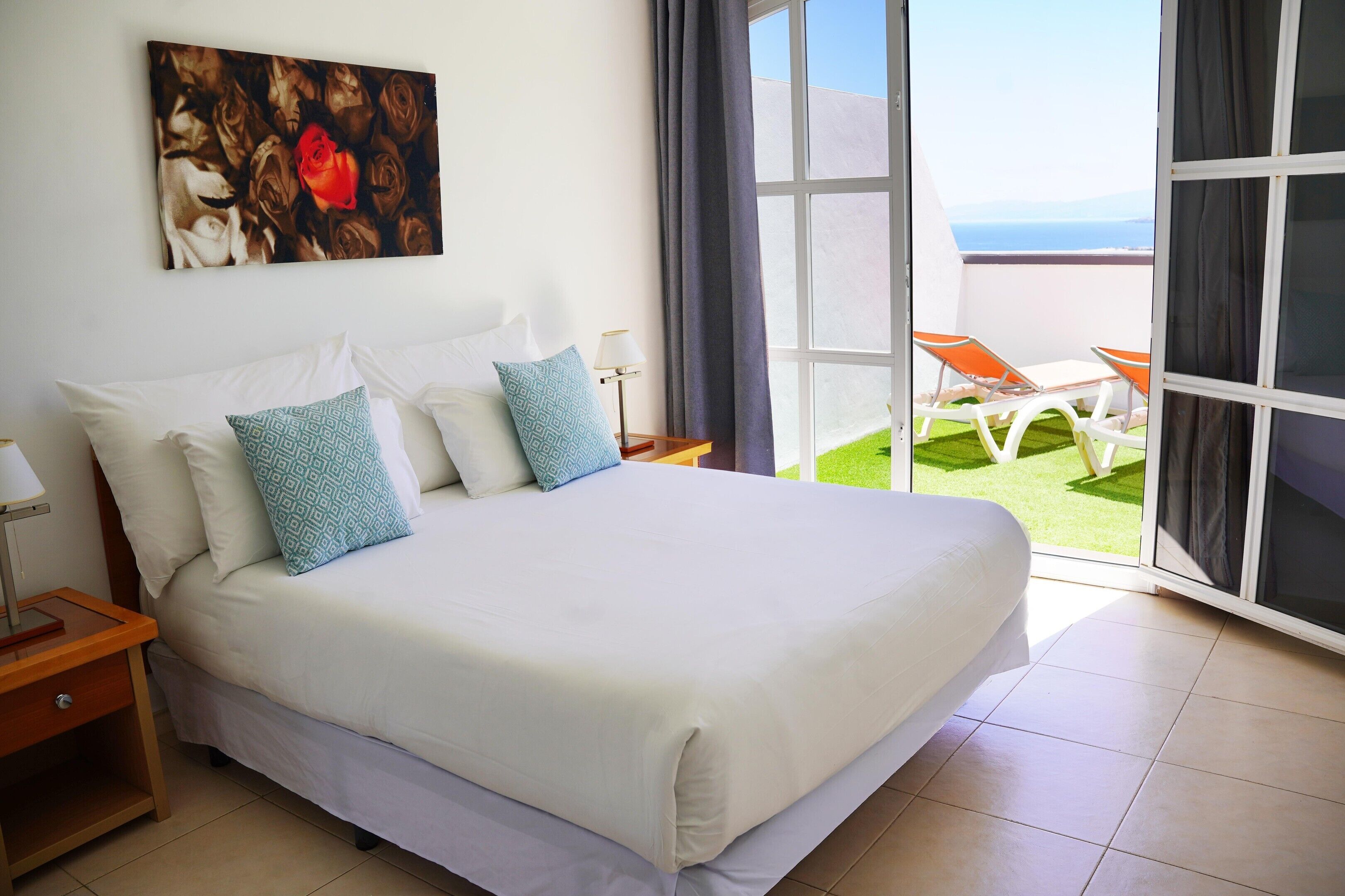 Quarto com cama, almofadas, mesa de cabeceira, um quadro e vista para uma varanda com cadeiras.