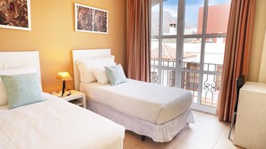 Quarto de hotel com duas camas, uma mesa de cabeceira com abajur, uma janela com cortinas e vista para prédios.