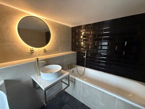 Uma casa de banho moderna com um espelho redondo, uma lavatório tipo cuba branco, uma banheira e uma parede revestida a mosaico preto.