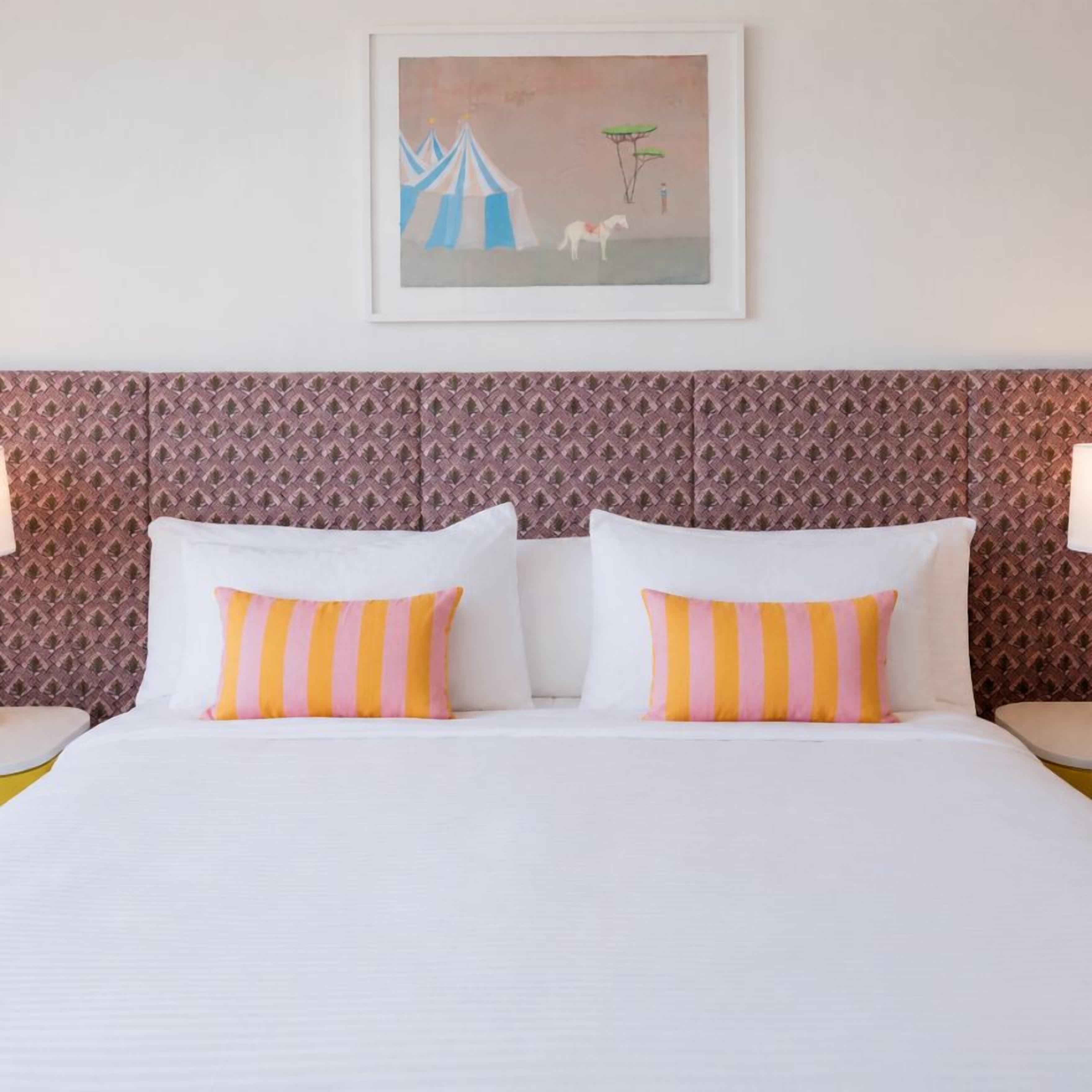 Uma cama de casal com roupa de cama branca e duas almofadas listradas, um painel de cabeceira estampado e um quadro emoldurado na parede.