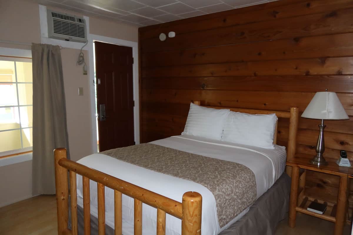 Quarto com painéis de madeira, uma cama, mesa de cabeceira, luminária e janela com cortinas.