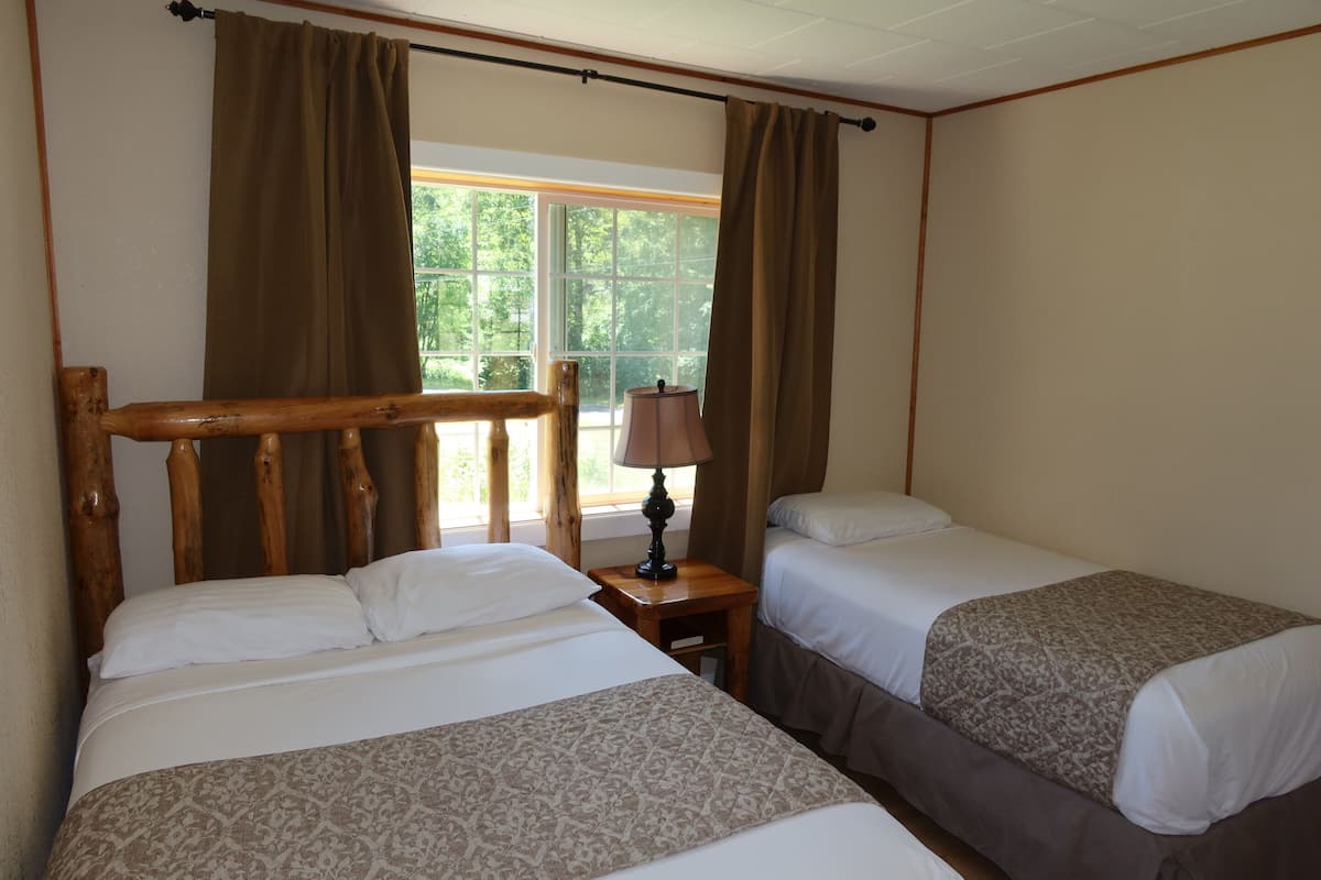 Quarto com duas camas, cabeceira de madeira, mesa de cabeceira com abajur e janela com cortinas.