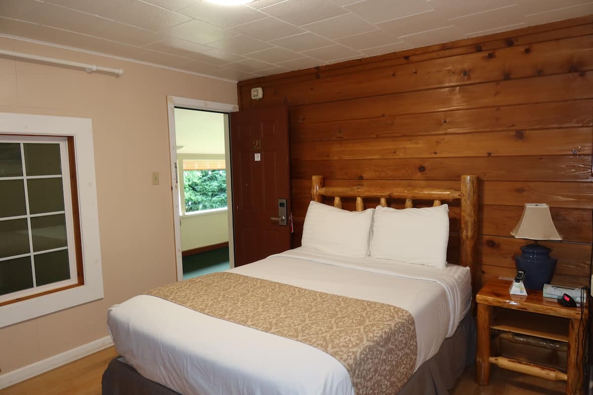 Quarto com cabeceira de madeira, cama com roupa de cama branca, mesa de cabeceira com abajur e porta que dá para uma varanda.