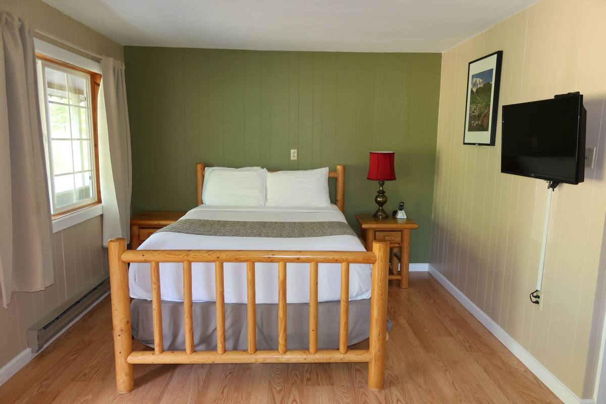 Quarto com cama de madeira, mesa de cabeceira, abajur vermelho, televisão e janela com cortinas.
