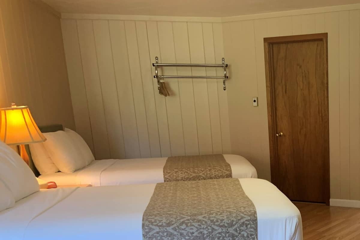 Quarto de hotel com duas camas, uma porta de madeira, um porta-toalhas fixado na parede e um abajur.