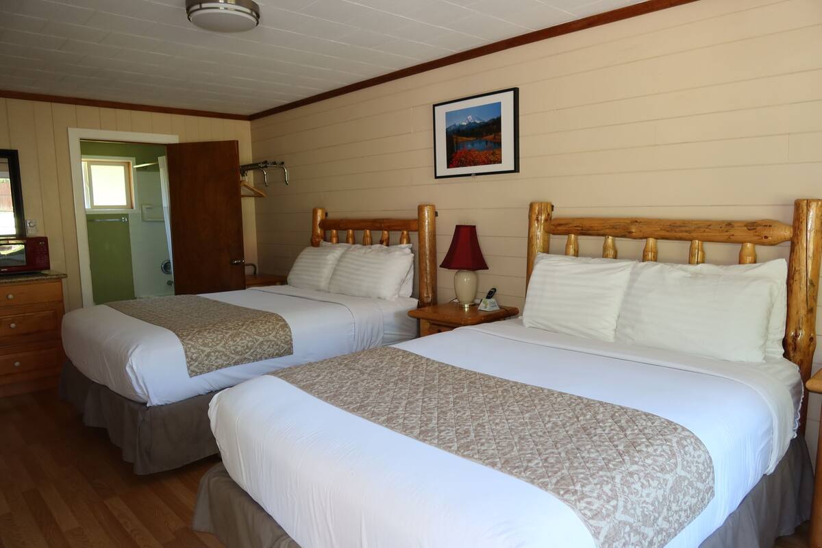 Quarto com duas camas, cabeceiras de madeira, mesa de cabeceira com abajur, um quadro na parede e uma porta que dá para outro cômodo.