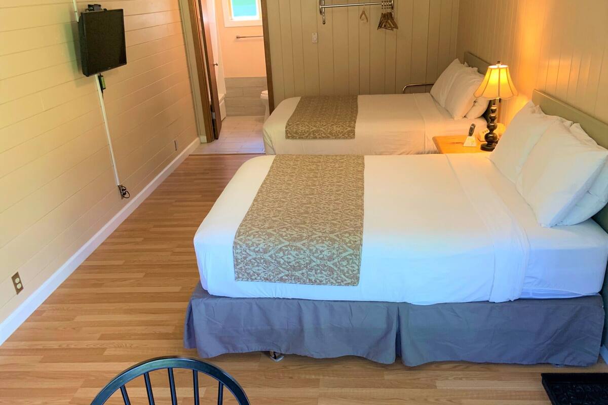 Quarto de hotel com duas camas, televisão fixada na parede, mesa de cabeceira com abajur e chão de madeira.