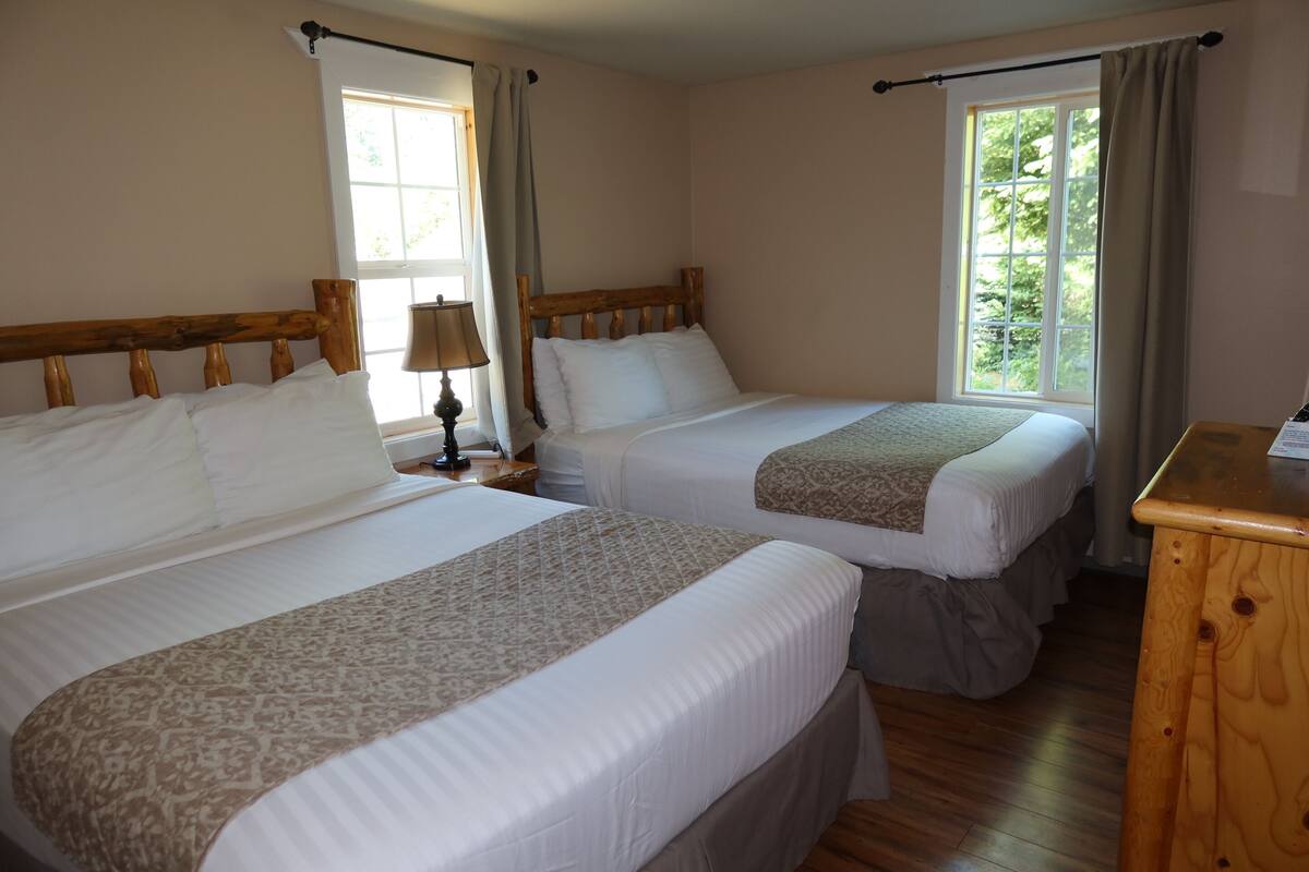 Quarto de hotel com duas camas, uma cabeceira de madeira, uma mesa de cabeceira com abajur e uma janela com cortinas.
