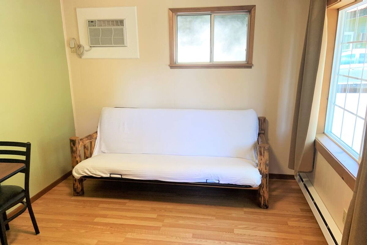 Quarto com piso de madeira, sofá branco, cadeira de madeira, janela e ar condicionado fixado na parede.