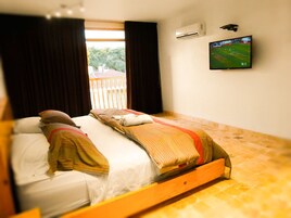 Cama de solteiro com estrutura de madeira, roupa de cama branca e manta bege. Há uma janela com cortinas escuras, um aparelho de ar condicionado na parede e uma televisão exibindo uma partida esportiva.