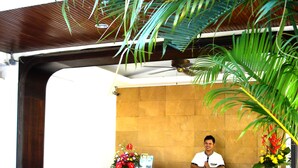 Uma área de recepção com recepcionista, mobiliário de madeira, balcão de atendimento e plantas tropicais.