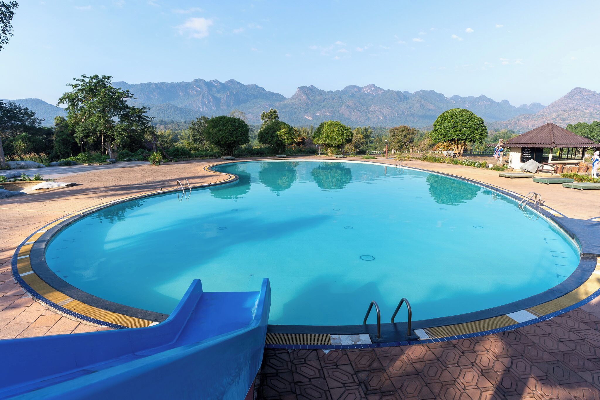 Piscina com escorrega, rodeada por área pavimentada e árvores, com montanhas ao fundo.