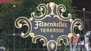 Uma placa com o texto "Felsenkeller TERRASSE" em fonte decorativa, fixada numa grade de metal com detalhes ornamentais.
