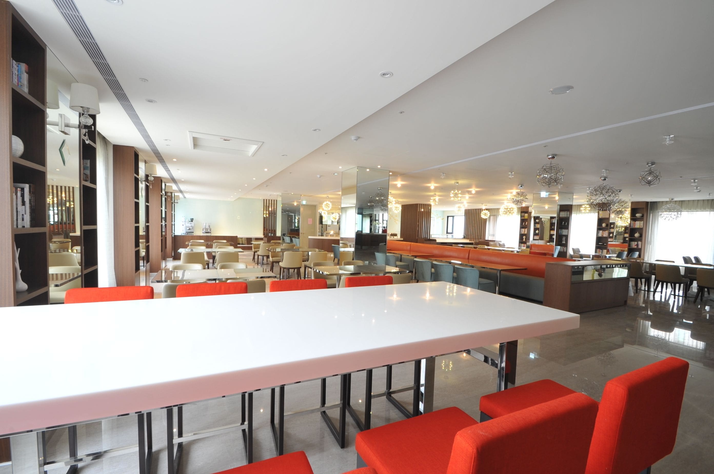 Amplo espaço interior com uma longa mesa branca rodeada por cadeiras laranja, zona de bar com assentos laranja e design moderno com pavimento e luminárias de teto refletoras.