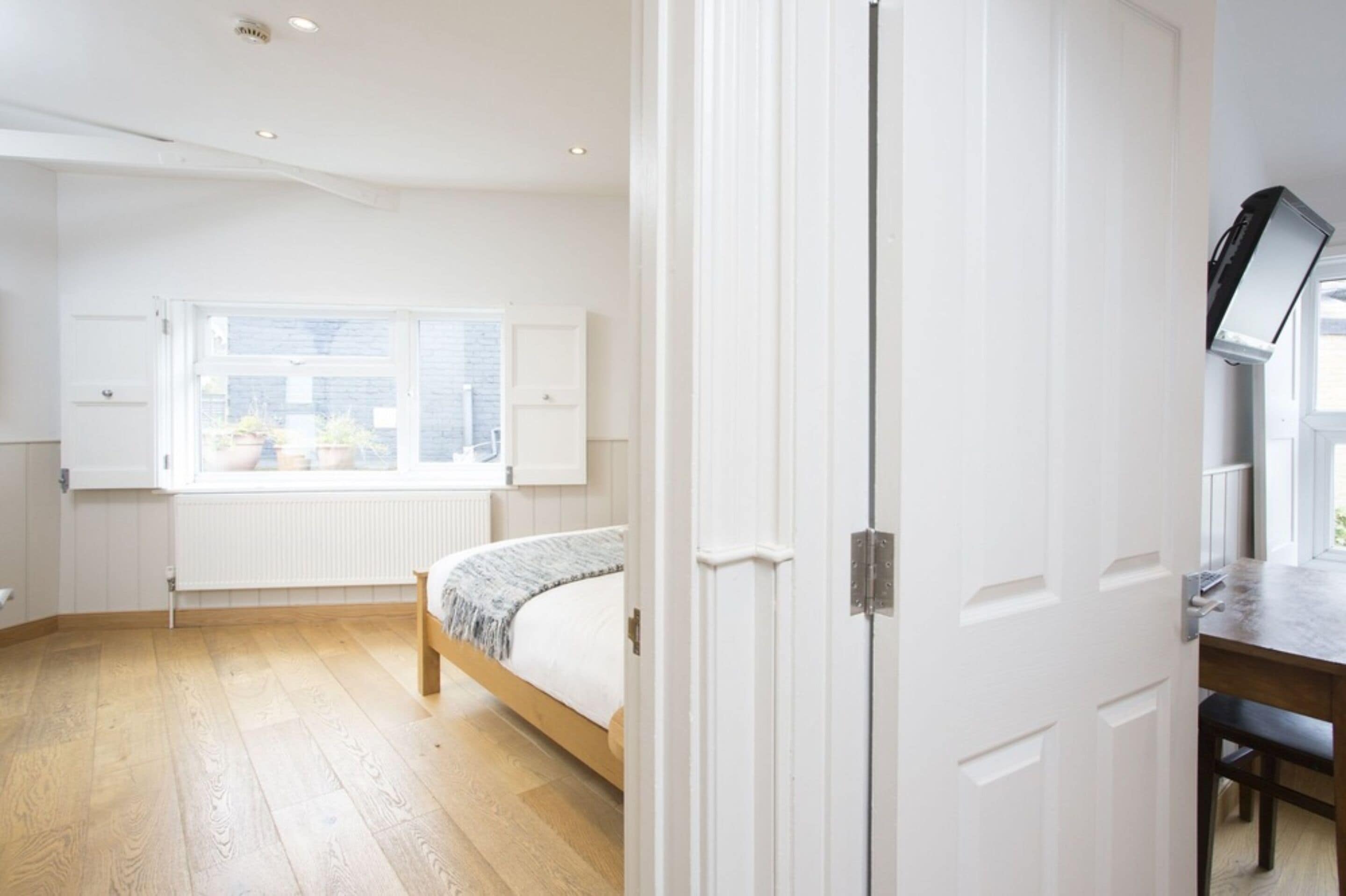 Quarto com piso de madeira, cama com cobertor cinza, janela com persianas brancas e radiador branco.