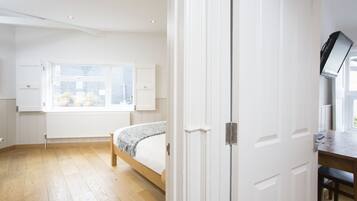 Quarto com piso de madeira, cama com cobertor cinza, janela com persianas brancas e radiador branco.