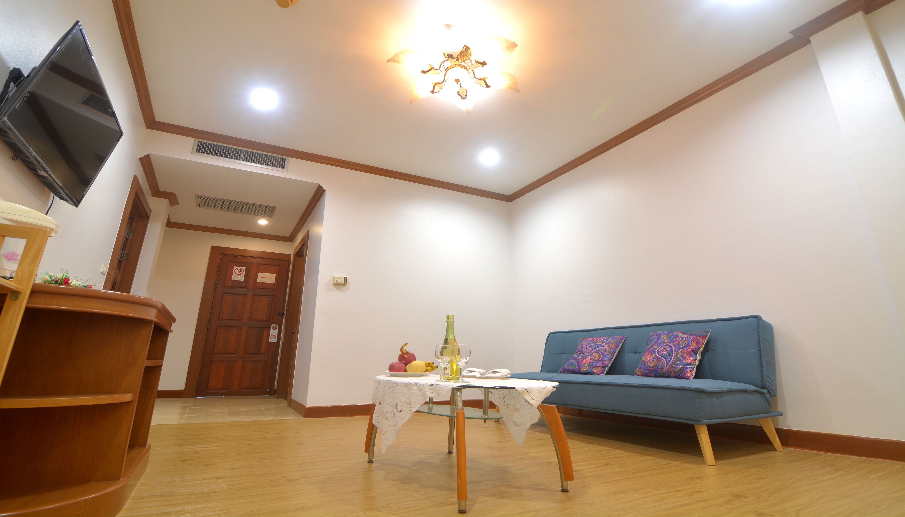 Um quarto com um sofá azul, uma mesa de madeira com toalha rendada, uma televisão na parede e uma escada de madeira que dá para outro cômodo.