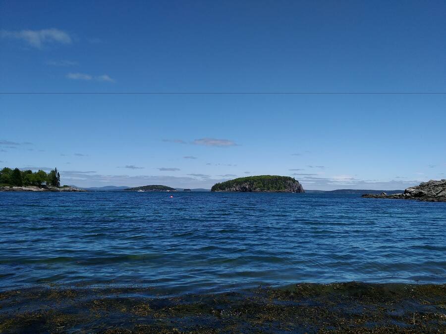 Paisagem costeira com águas calmas, uma pequena ilha com vegetação e montanhas distantes sob um céu claro.