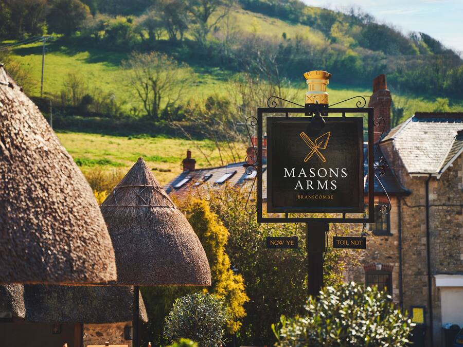 Placa do Masons Arms em Bancombe, com telhado de palha e vista para colinas verdes ao fundo.