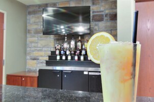 Uma área de bar com parede de pedra, uma TV de tela plana montada acima de uma fileira de garrafas de bebidas alcoólicas e um copo com uma fatia de limão no balcão.