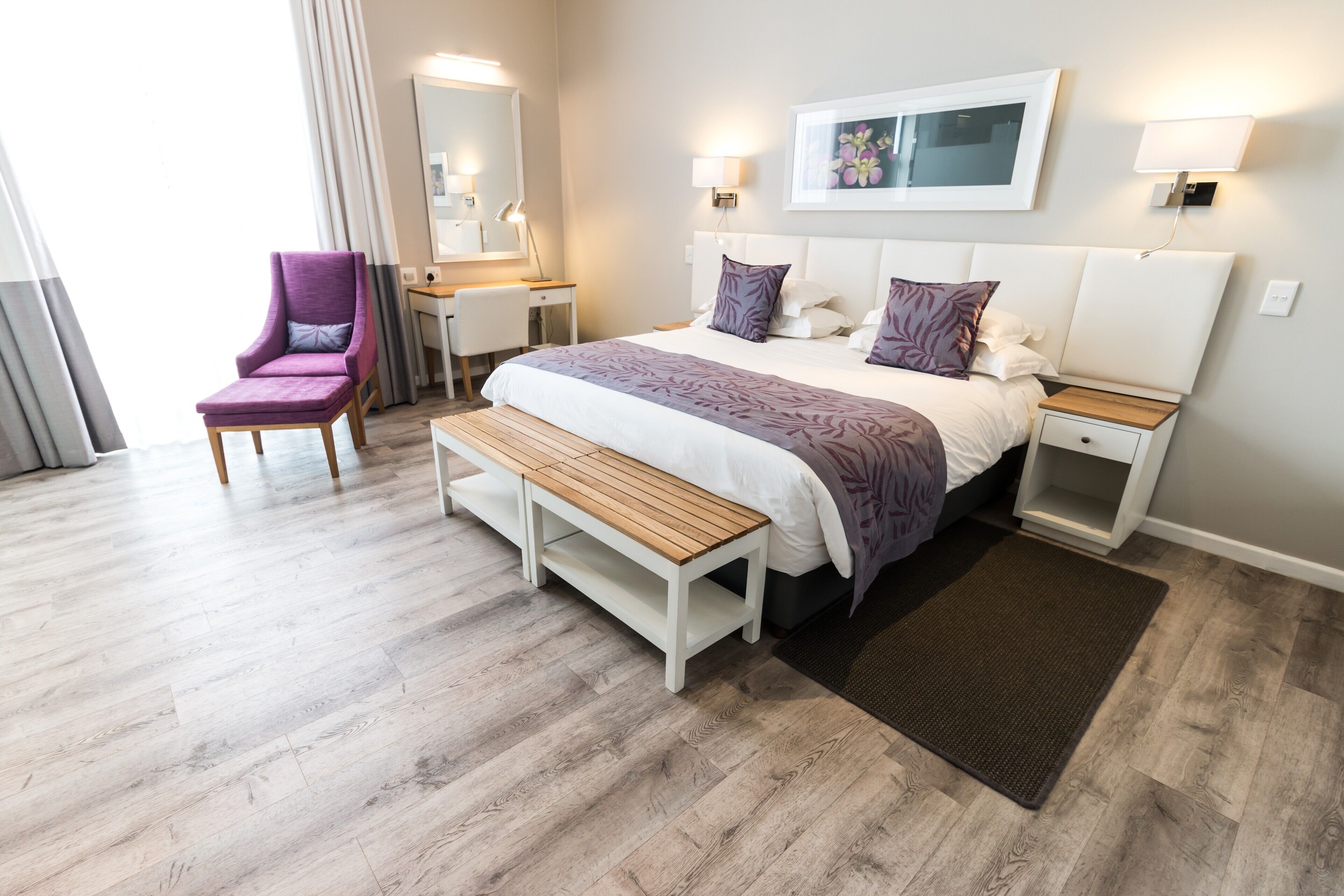 Quarto de hotel moderno com uma cama grande, um banco de madeira, uma escrivaninha e uma poltrona roxa.