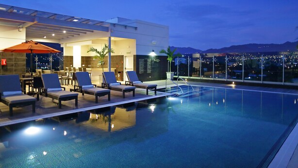 Piscina infinita no terraço com espreguiçadeiras e área de refeições, com vista para a paisagem urbana ao entardecer.
