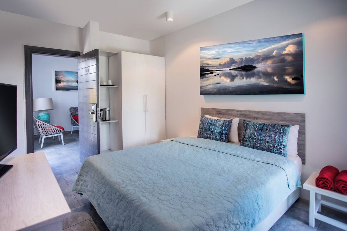 Quarto de hotel moderno com cama, televisão, cadeira e pintura de um pôr do sol sobre a água.