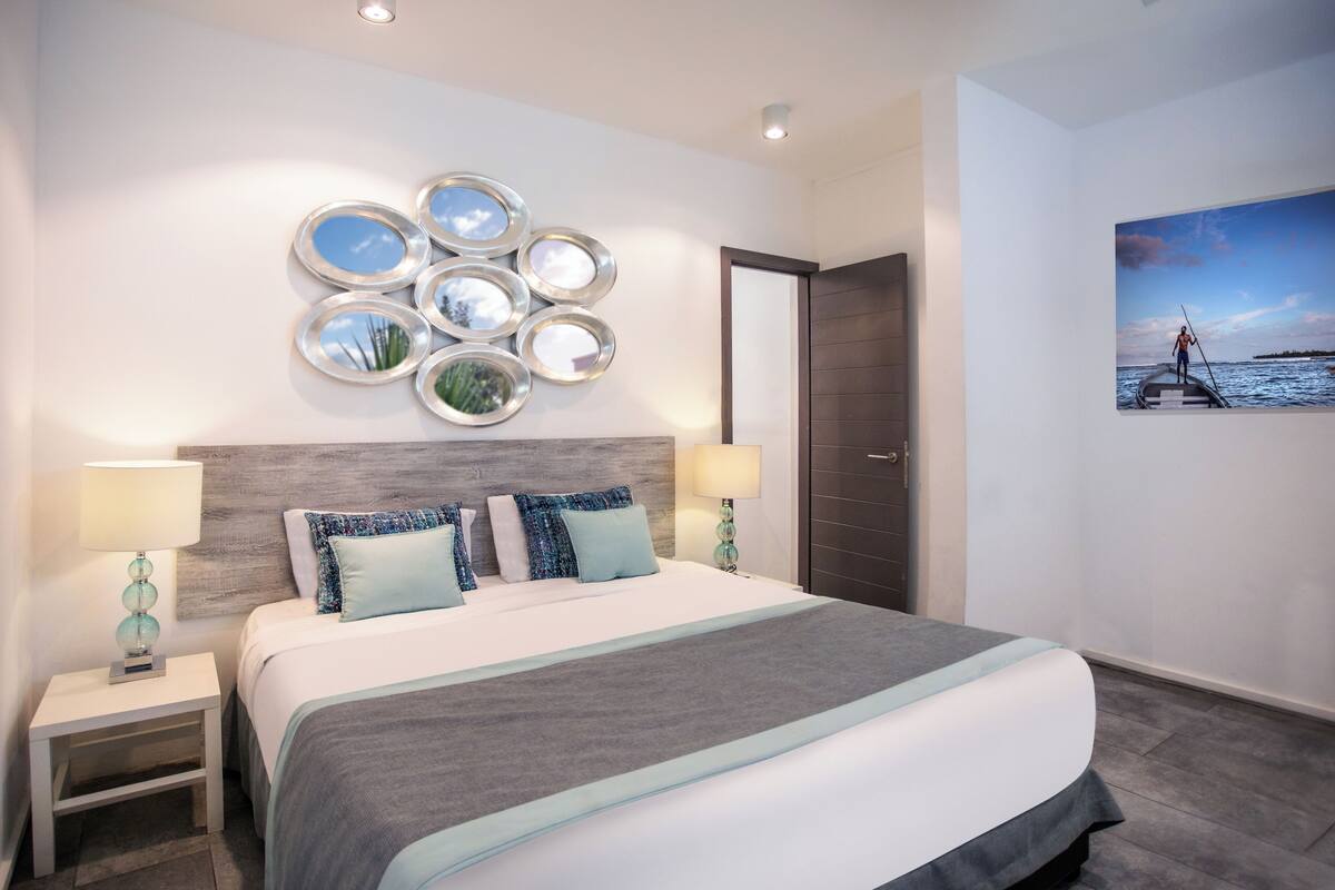 Quarto de hotel moderno com uma cama grande, duas mesas de cabeceira com luminárias, um arranjo de espelhos fixados na parede e um quadro com uma imagem de um barco na água.