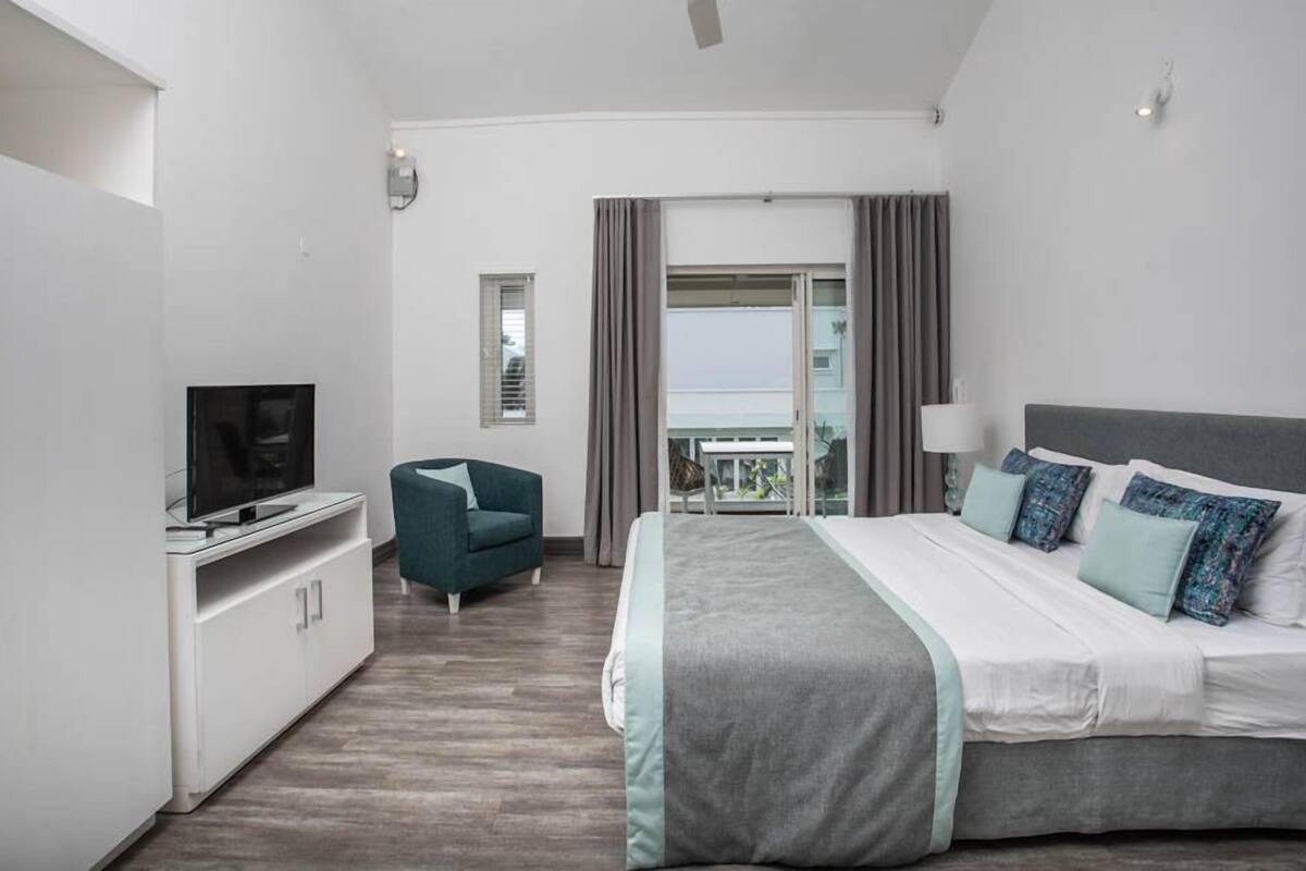 Quarto de hotel moderno com uma cama grande, uma televisão sobre um móvel branco, uma poltrona azul-turquesa e uma varanda com cortinas.