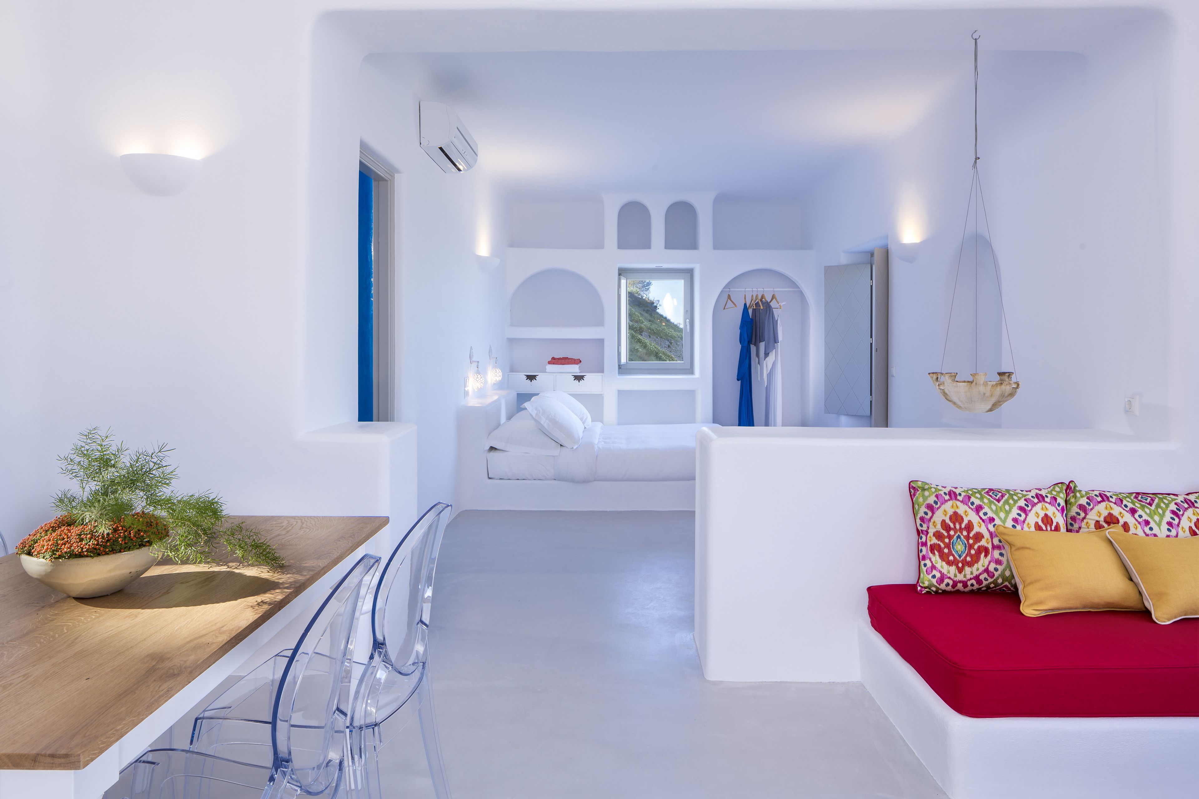 Quarto moderno e minimalista com mesa de jantar de madeira, cadeiras de plástico transparente, sofá vermelho e cama branca com vista para a janela.