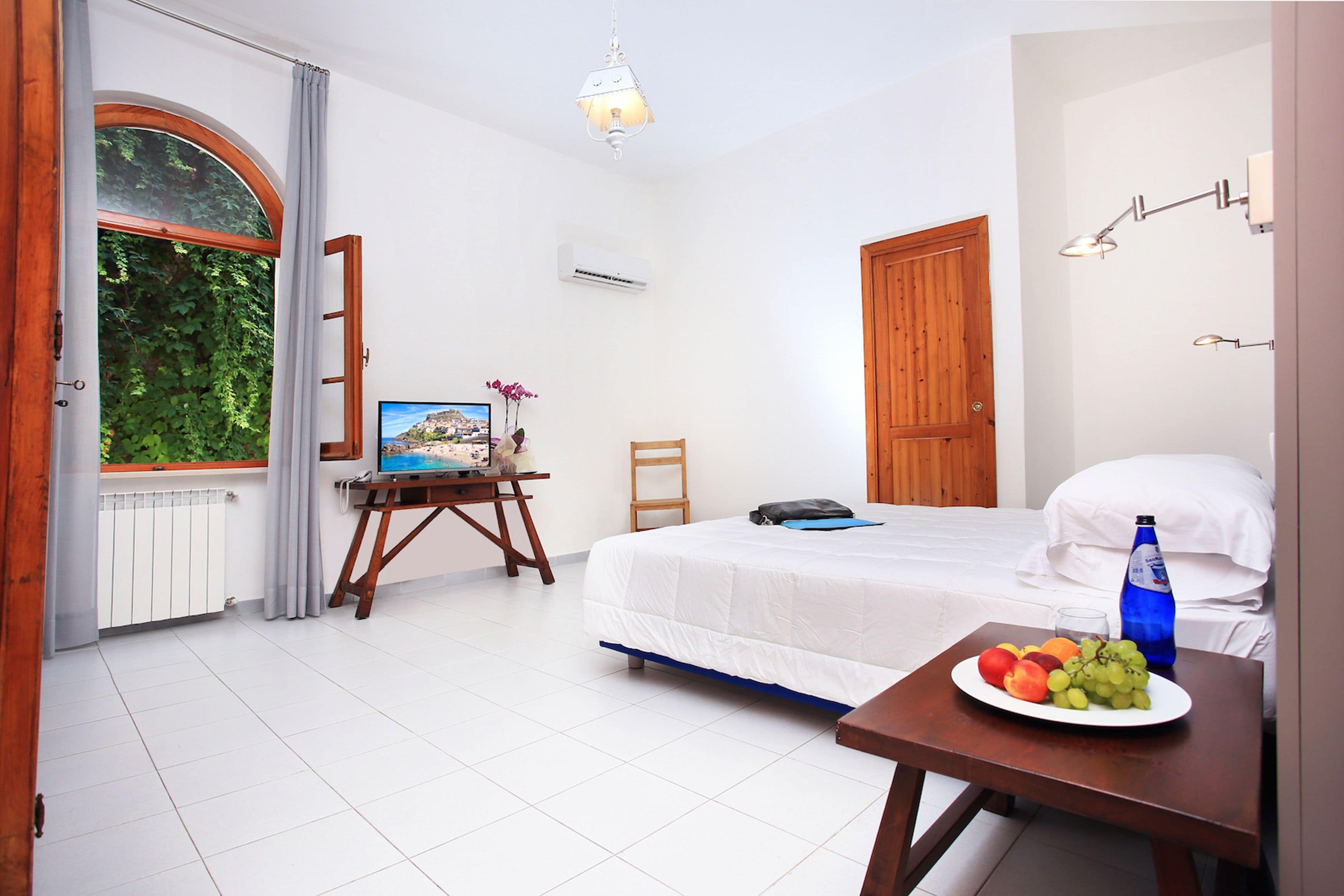 Quarto de hotel com uma cama, uma mesa com uma travessa de fruta, uma pintura em um cavalete e vista para o verde pela janela.