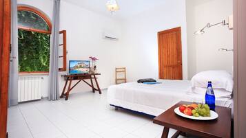 Quarto de hotel com uma cama, uma mesa com uma travessa de fruta, uma pintura em um cavalete e vista para o verde pela janela.