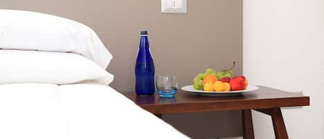 Uma cama bem arrumada com lençóis brancos, uma mesa de cabeceira de madeira com um frasco azul, um copo e um prato de frutas.