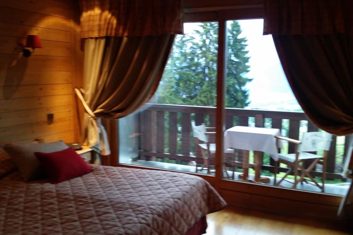 Quarto com cama, janela com cortinas, vista para o exterior e varanda com mesa e cadeiras.