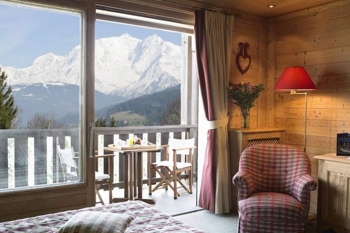 Quarto com uma cama, uma poltrona xadrez, uma pequena mesa com cadeiras, um abajur vermelho e vista para montanhas nevadas.