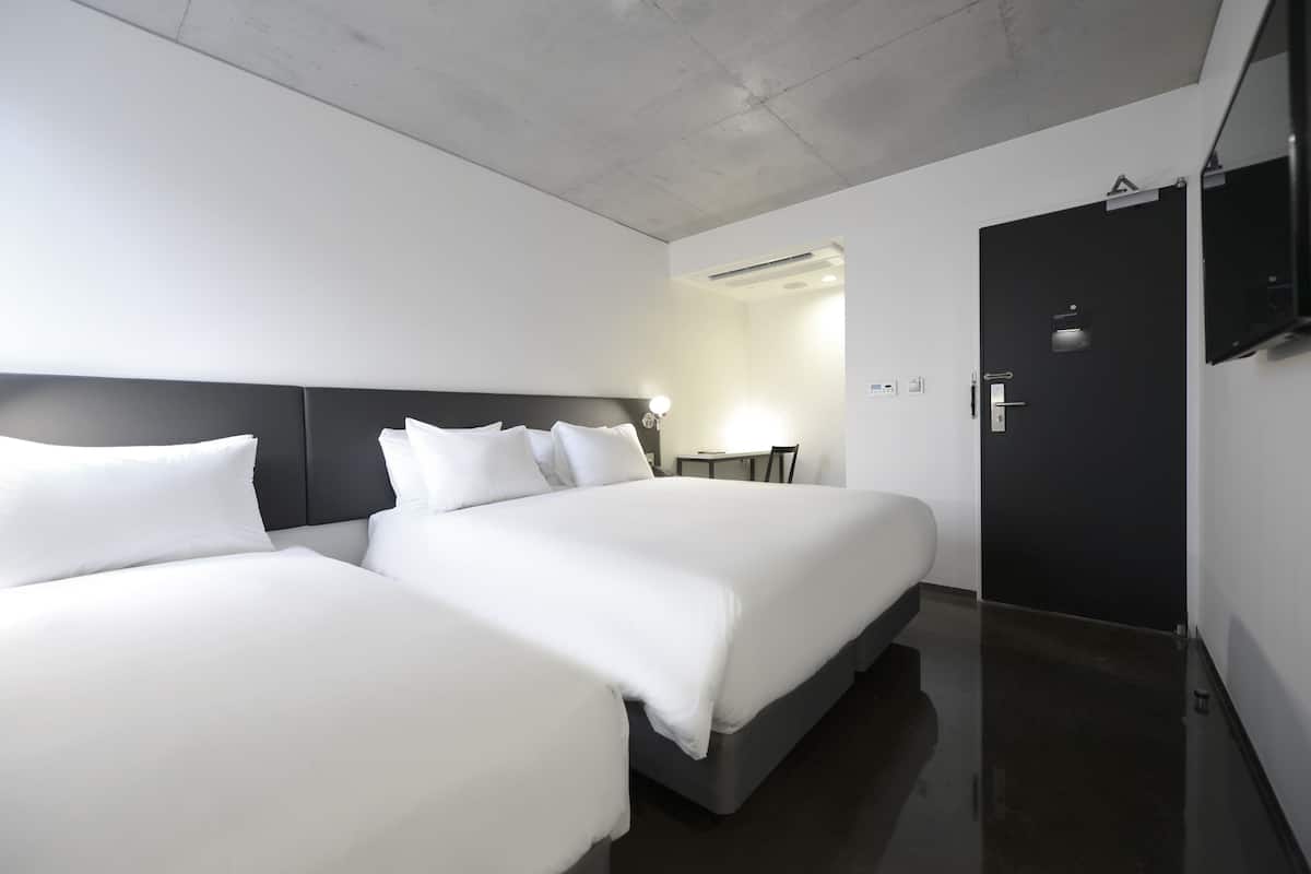 Quarto de hotel moderno com duas camas, uma porta escura e uma TV de tela plana fixada na parede.