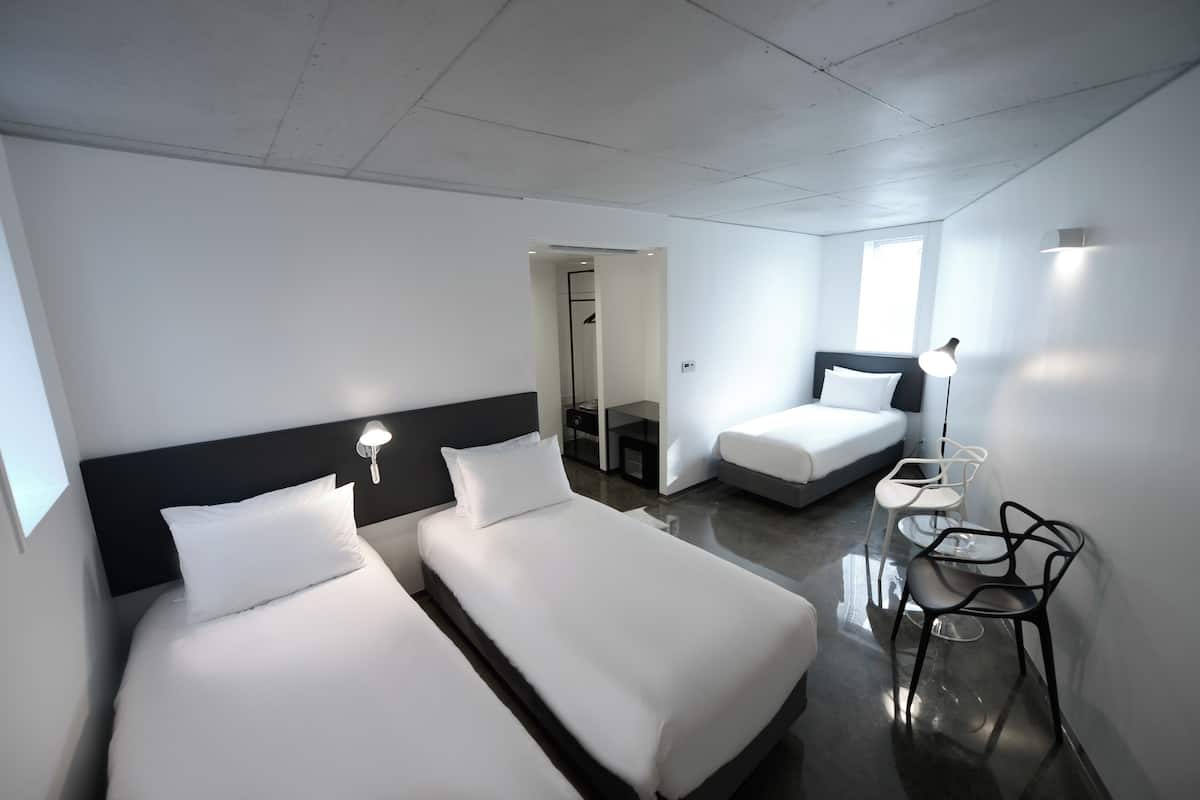 Quarto de hotel moderno com duas camas de solteiro, uma mesinha e uma cadeira.